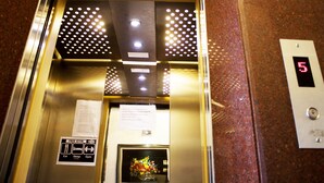 Interior de elevador com visor digital exibindo o número 5, placa indicando acesso gratuito à academia e pequena janela com arranjo de flores.
