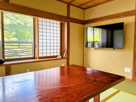 Um quarto japonês tradicional com tatames, uma mesa de madeira, uma TV e uma janela com vista para um deck de madeira e vegetação.