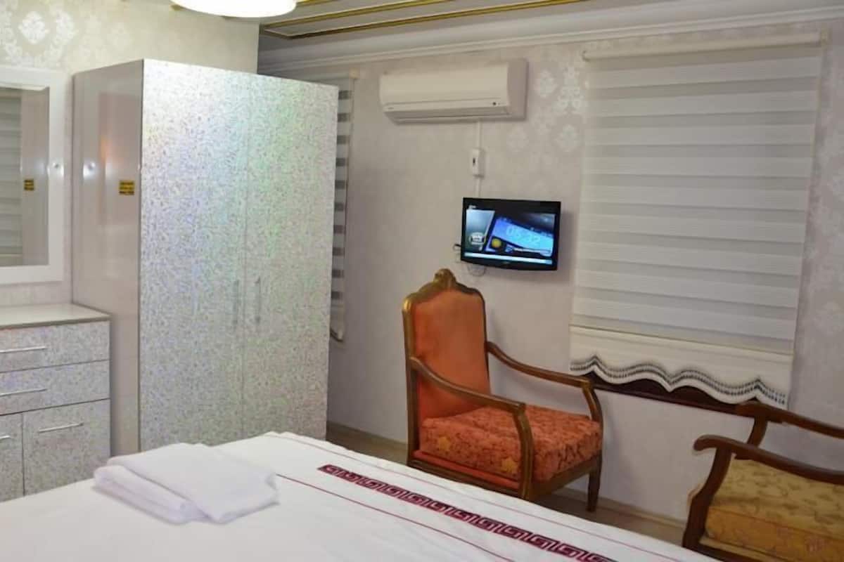 Quarto de hotel com cama, guarda-roupa, cadeira, televisão e janela com persianas.