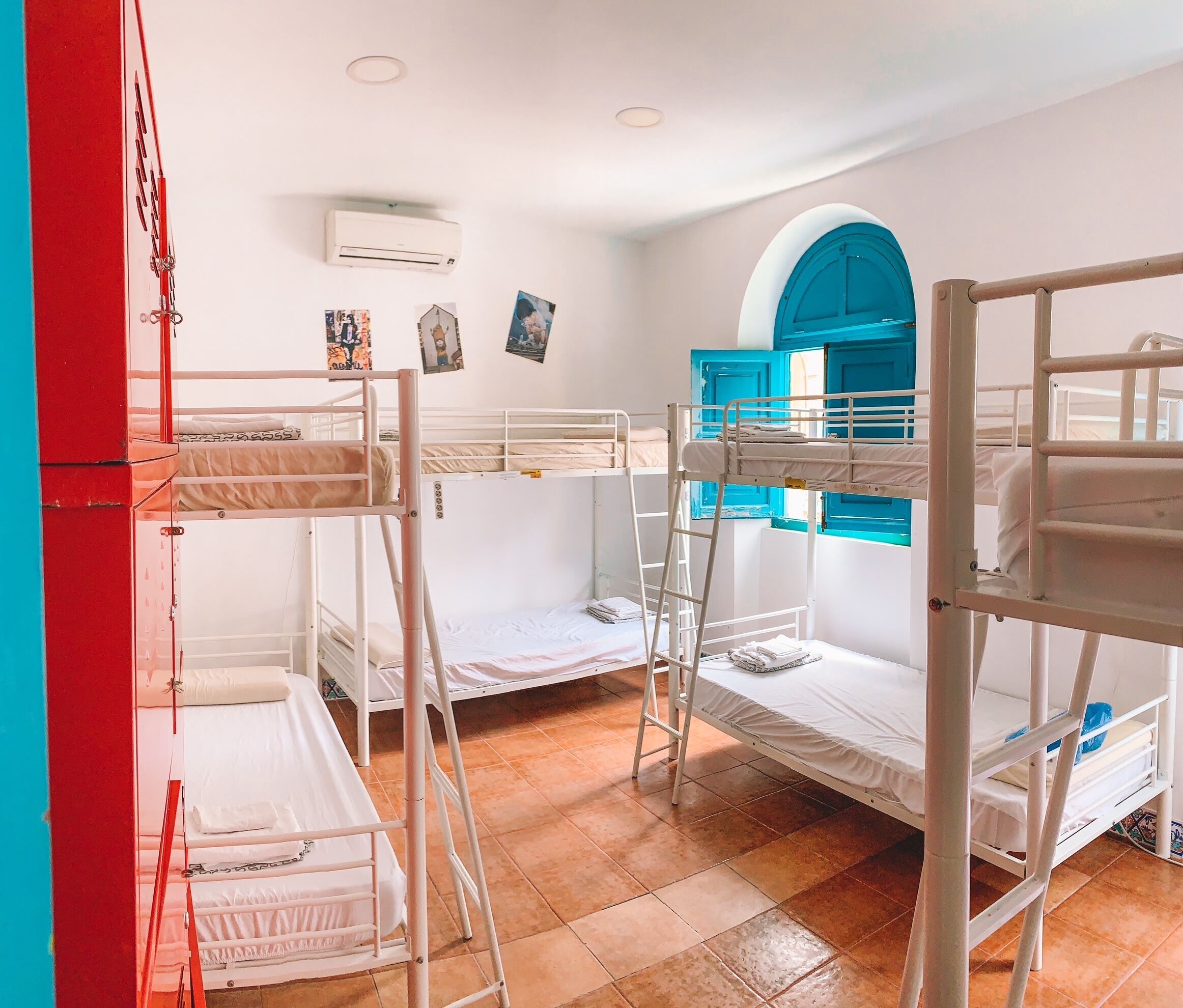 Quarto de estudante com beliches, ar condicionado e janela com persianas azuis.