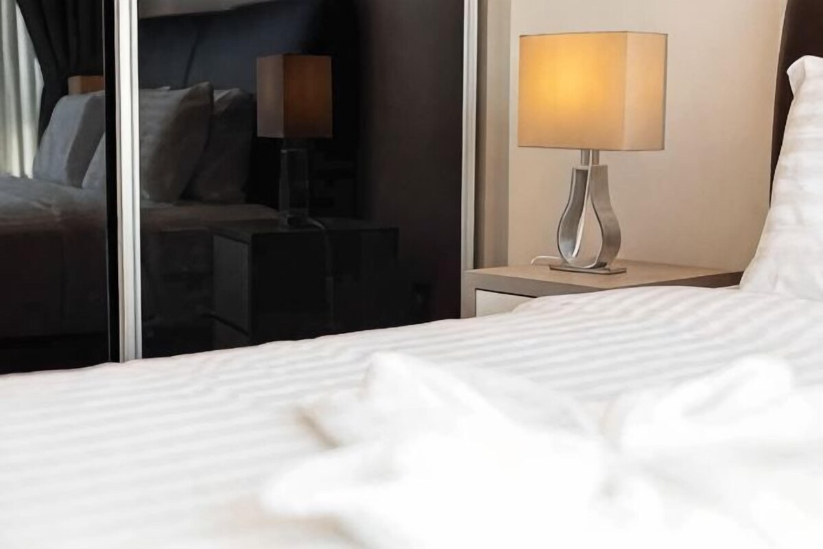 Quarto de hotel com cama, mesa de cabeceira, abajur e espelho refletindo o ambiente.