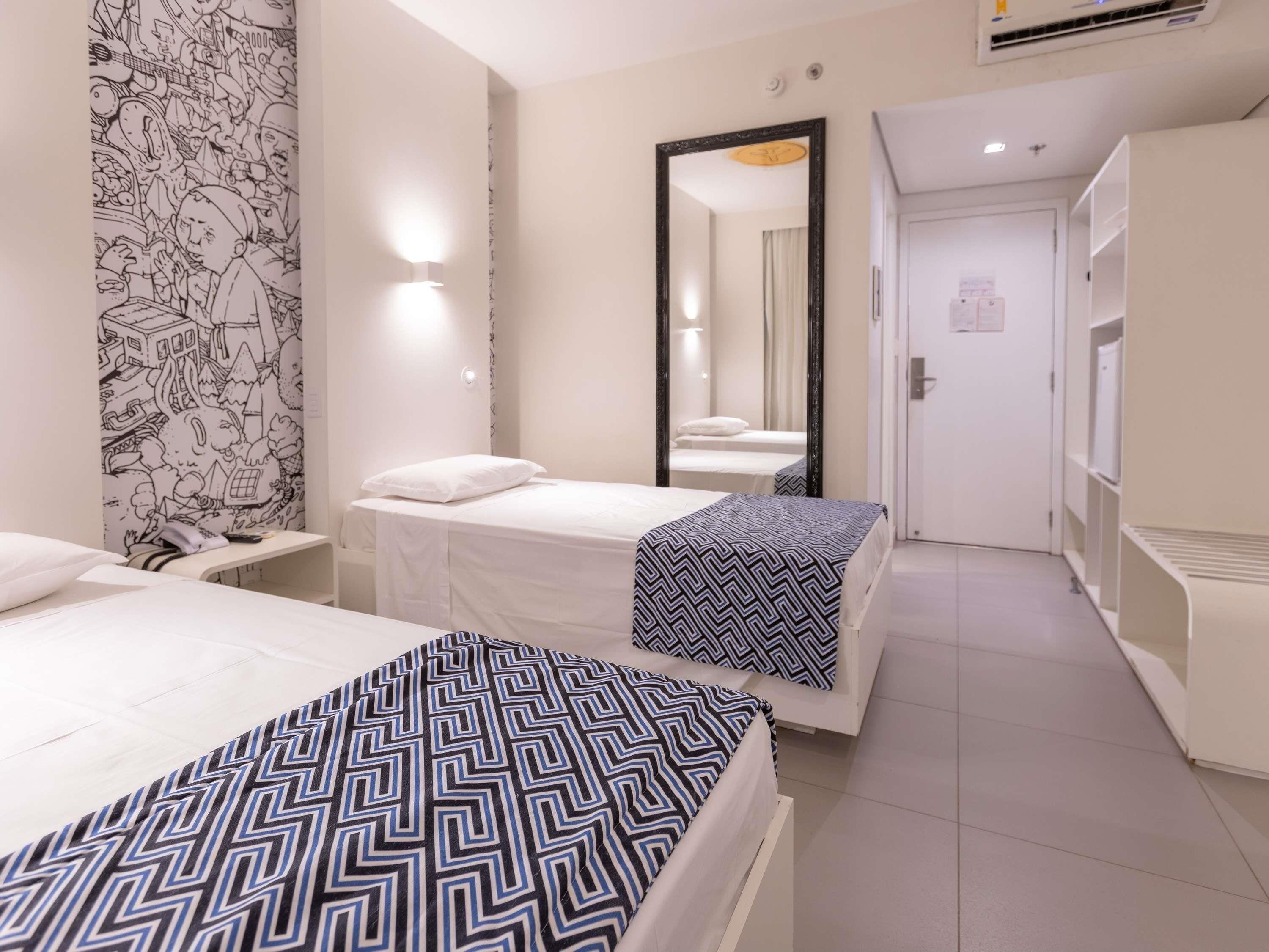 Quarto de hotel com duas camas, um espelho grande, um guarda-roupa e uma parede com padrão em preto e branco.