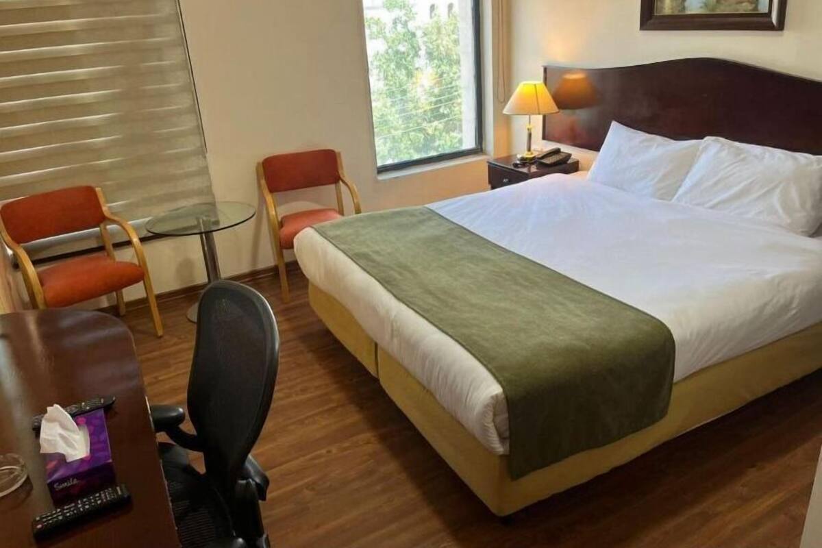 Quarto de hotel com uma cama grande, uma escrivaninha com cadeira, uma mesinha e uma janela com vista para o verde.