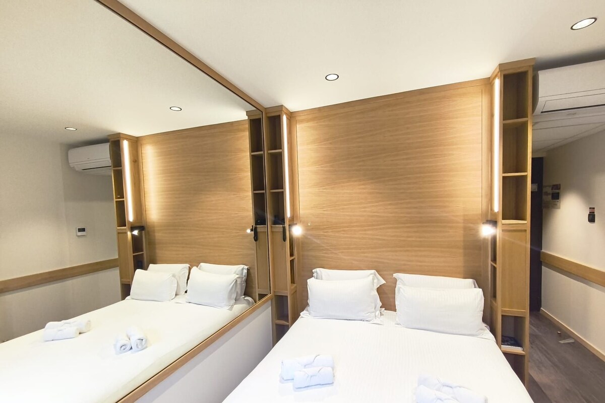 Quarto de hotel com duas camas, painéis de madeira e estantes embutidas.