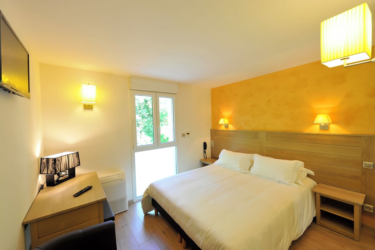 Quarto de hotel com cama, mesas de cabeceira, escrivaninha com luminária e janela.