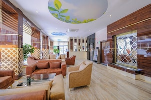 Um hall amplo com design moderno, apresentando uma grande área de estar com sofás e poltronas, teto decorativo com motivos de folhas e um grande janelão com moldura ornamental.
