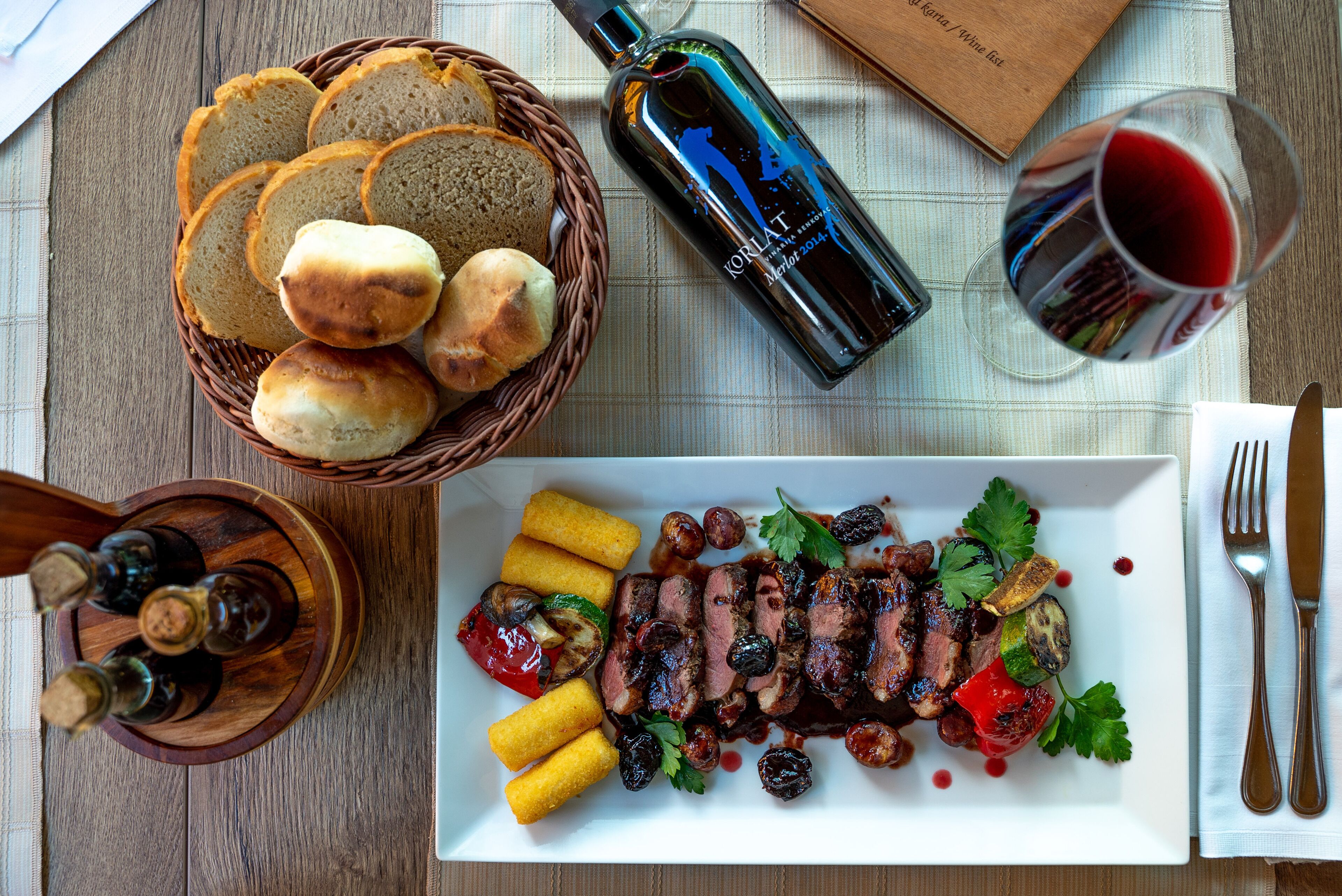 Uma mesa de jantar com um prato de bife, legumes e pães. Há também garrafas e um copo de vinho tinto.