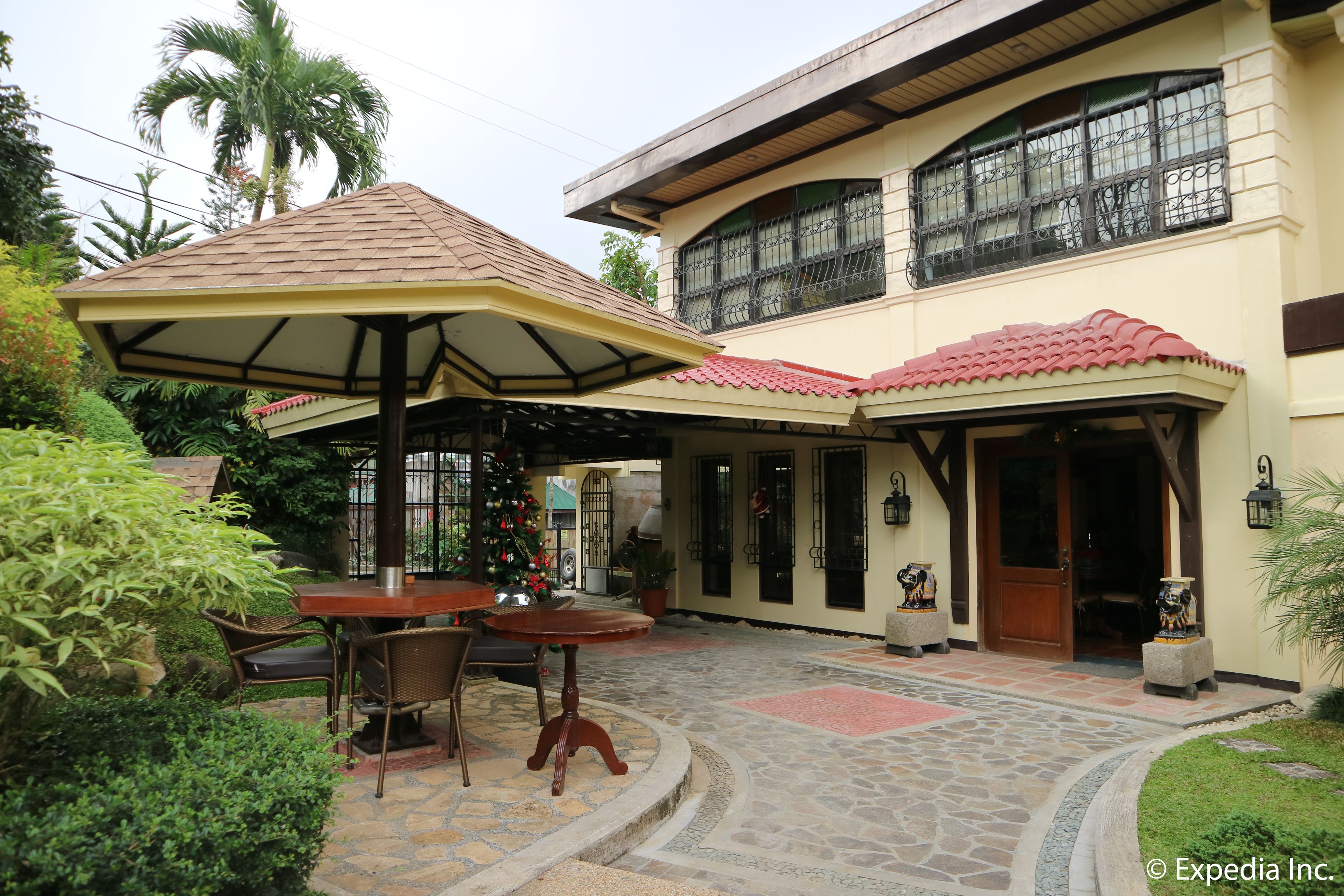 Uma área externa coberta com mesa e cadeiras de pátio, uma árvore de Natal e um edifício com telhado de telhas vermelhas e grandes janelas.