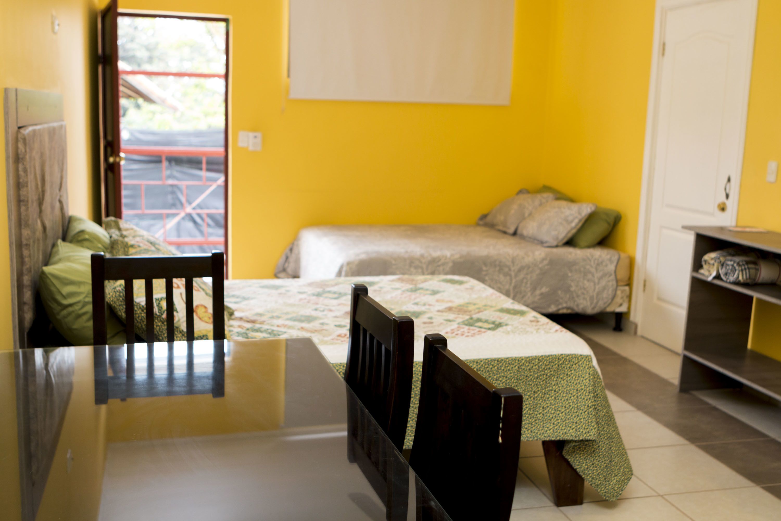Quarto com uma cama, uma cadeira e uma mesa. Tem uma porta e uma janela.