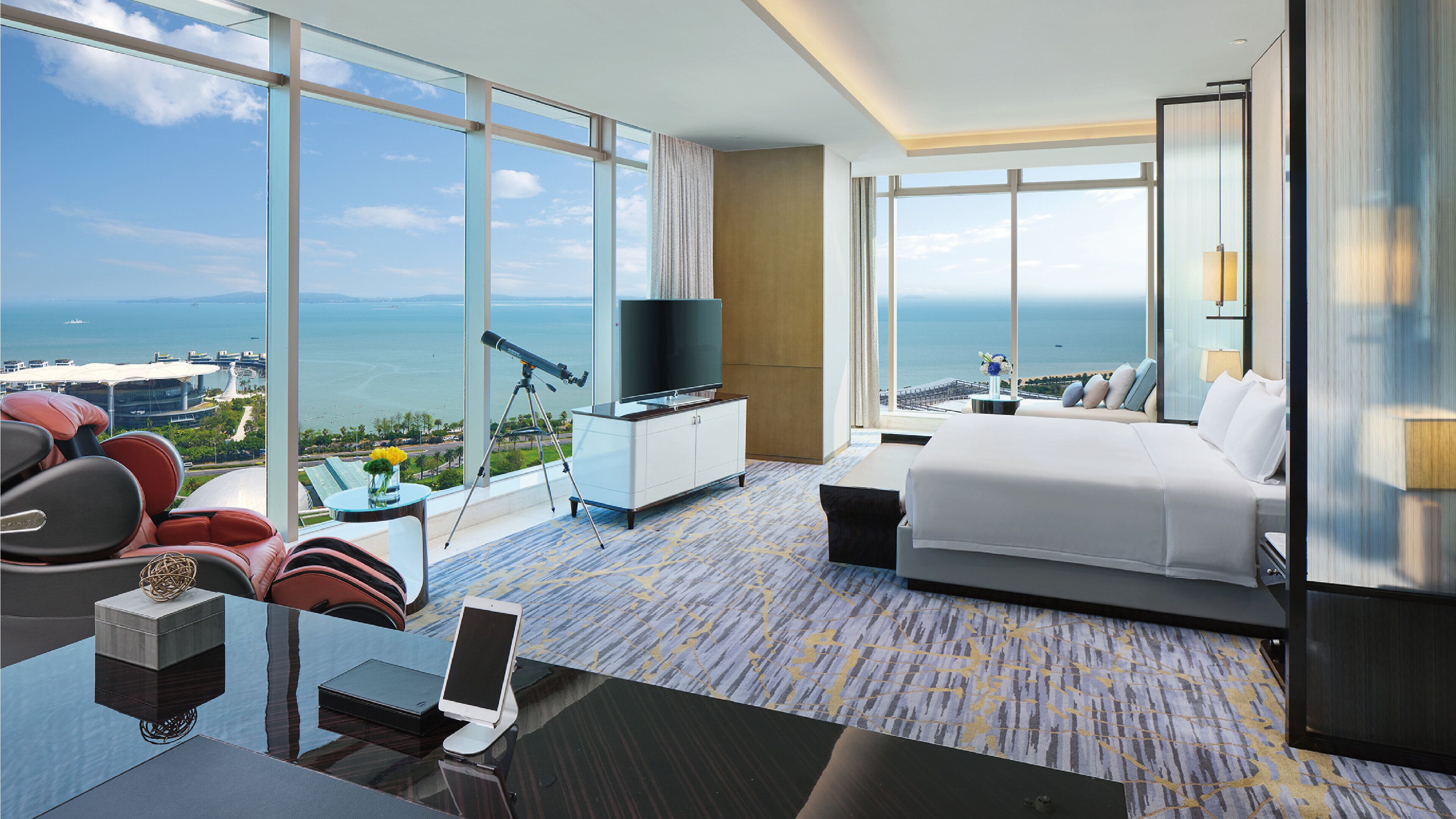 Quarto de hotel com uma janela grande, uma cama, uma escrivaninha com computador e um espaço de estar com vista para o mar.