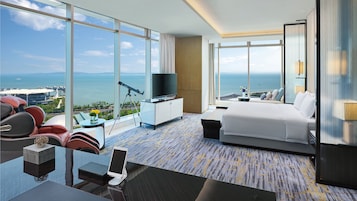 Quarto de hotel com uma janela grande, uma cama, uma escrivaninha com computador e um espaço de estar com vista para o mar.