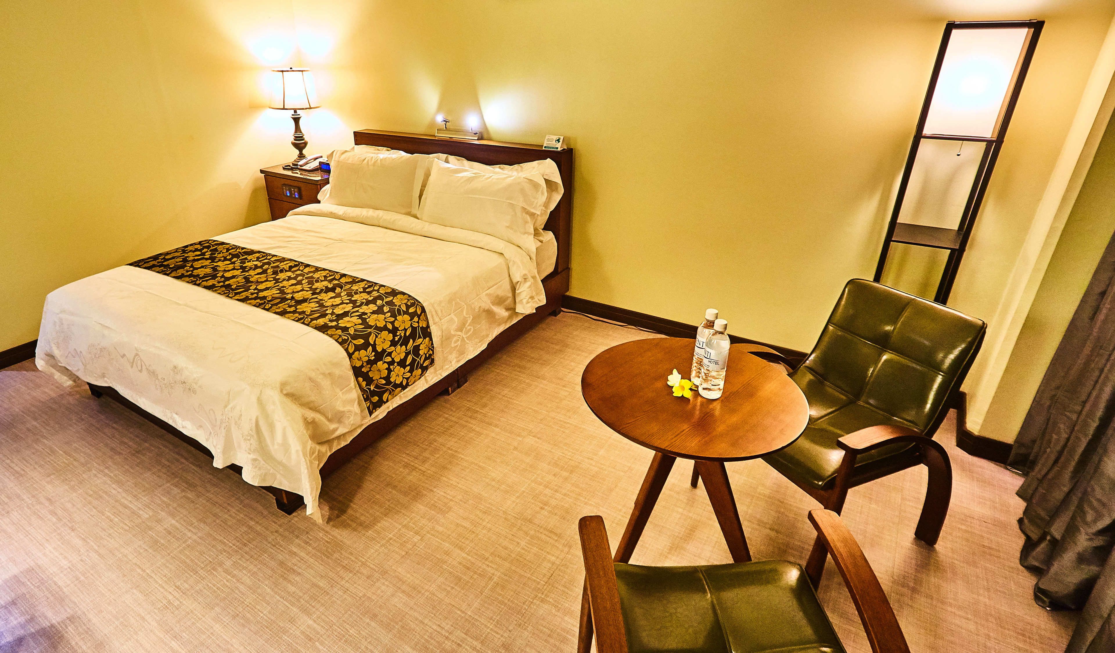 Quarto de hotel com cama, mesa de cabeceira com abajur, pequena mesa com garrafas de água e uma cadeira verde.