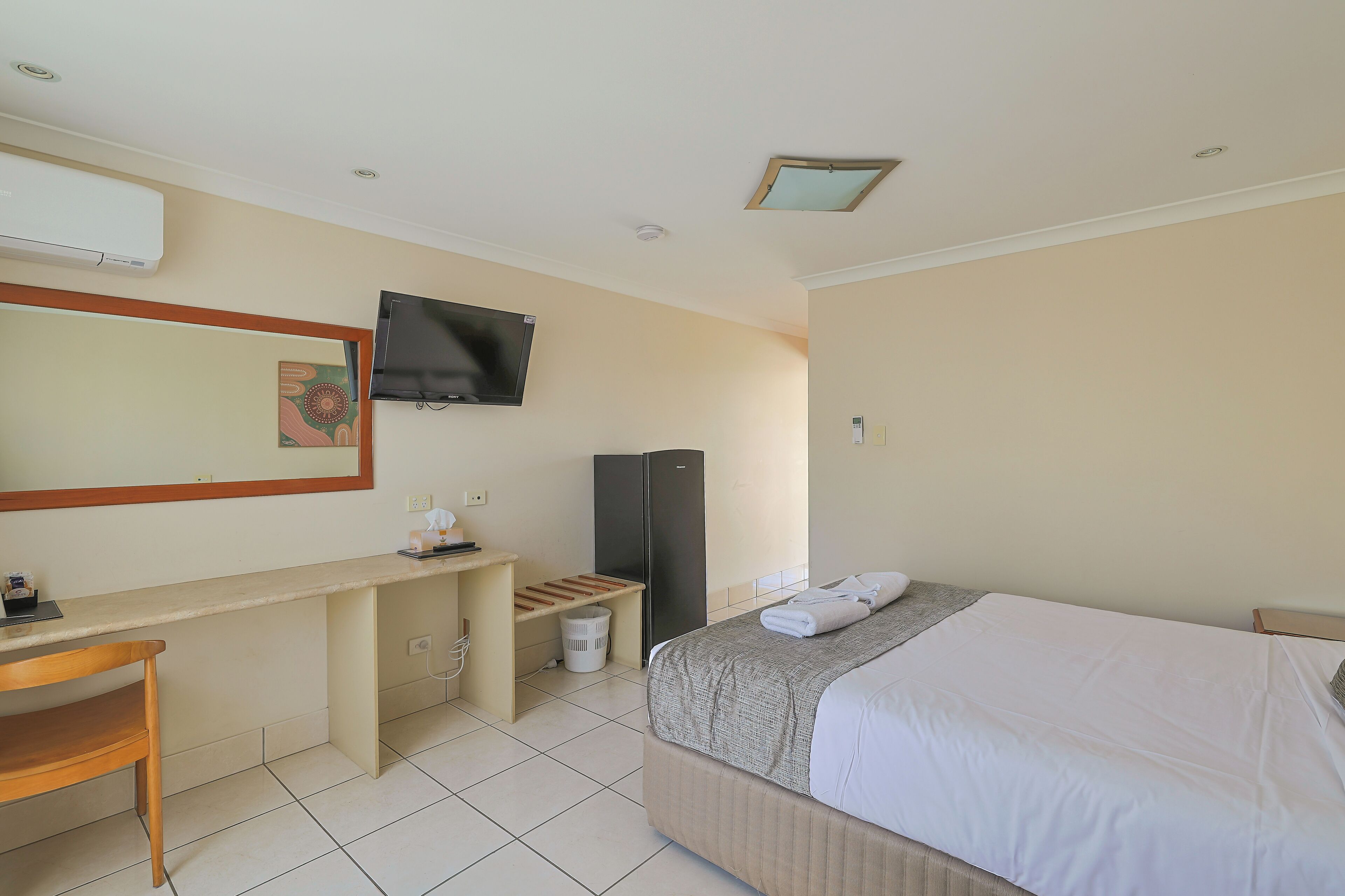 Quarto de hotel com cama, escrivaninha, cadeira, televisão e guarda-roupa.