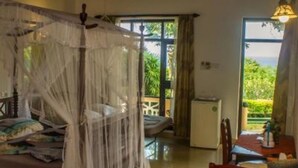 Quarto com cama de dossel, teto de madeira, área de jantar com mesa e cadeiras, e vista para o verde lá fora.