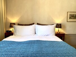 Uma cama bem arrumada com roupa de cama branca e azul, duas luminárias de cabeceira com cúpulas estampadas e um quadro na parede.