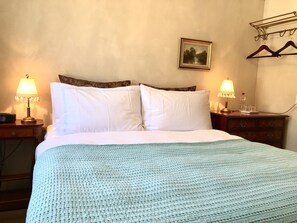 Cama bem arrumada com almofadas brancas e um cobertor azul, duas luminárias de cabeceira, um quadro na parede e um cabideiro com cabides.