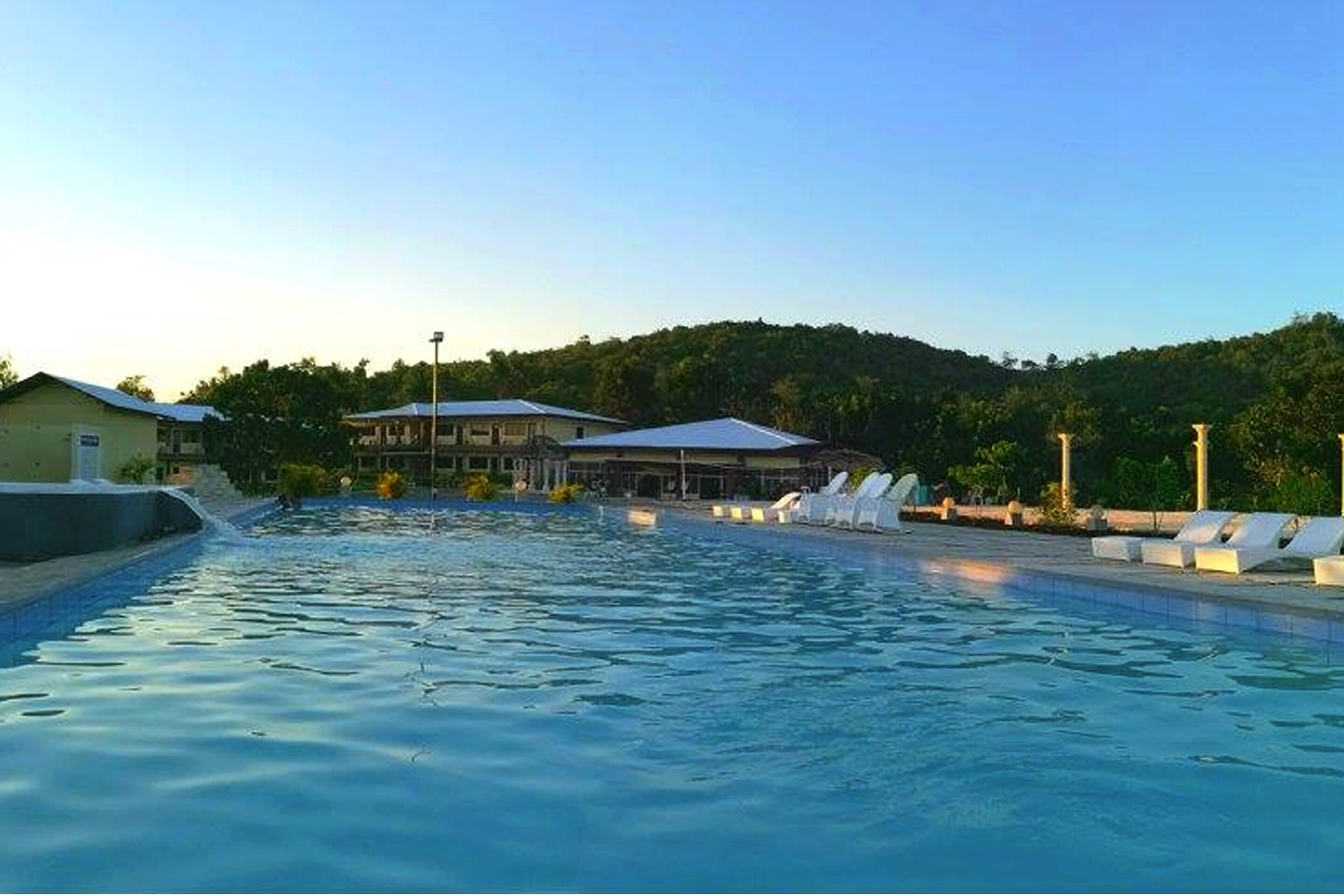 Uma piscina com espreguiçadeiras, um edifício com telhado plano e uma colina arborizada ao fundo.