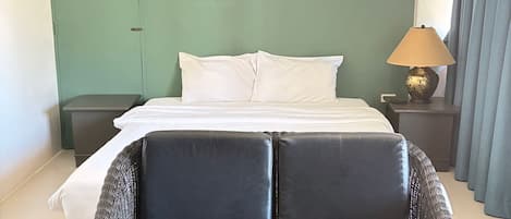 Quarto de hotel com cama, mesa de cabeceira, luminária, cadeira e porta.