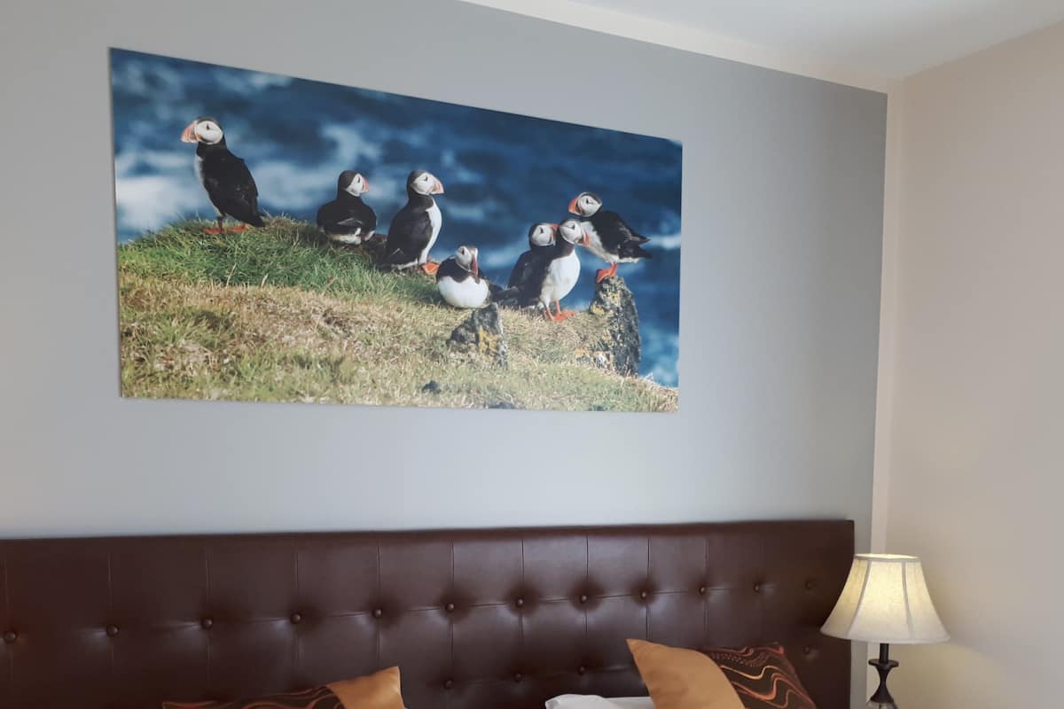 Quarto de hotel com uma cama grande, duas almofadas, mesa de cabeceira com abajur e pintura de pinguins na parede.