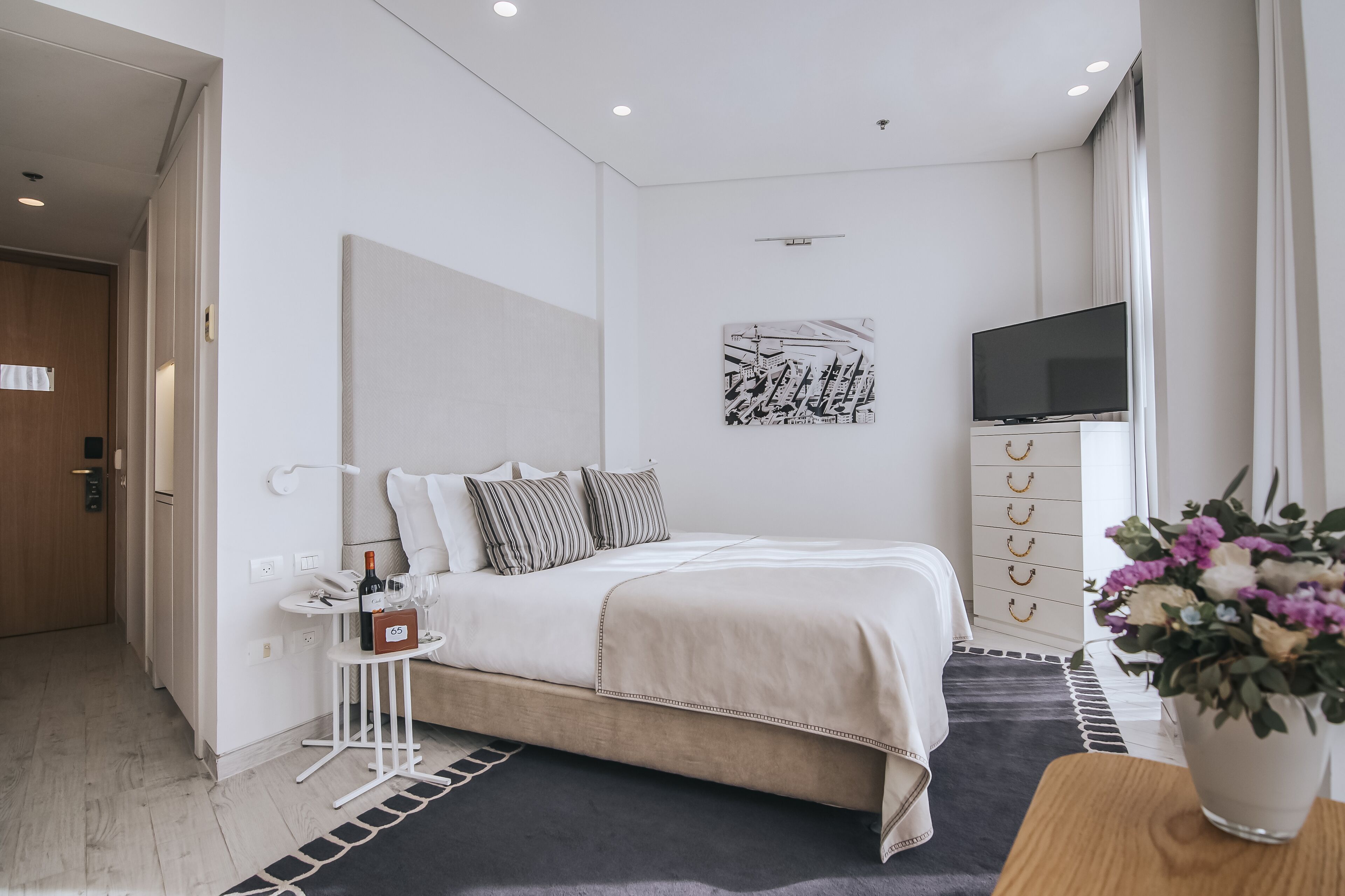 Quarto moderno com cama, televisão, uma mesinha com uma garrafa e um vaso com flores.