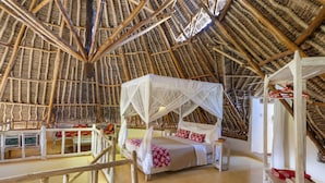 Um quarto com telhado de palha, uma cama com dossel e um banco de madeira.