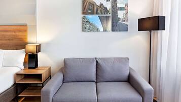 Quarto de hotel moderno com um sofá cinza, uma mesa de cabeceira de madeira com uma luminária, uma cama com lençóis brancos e uma obra de arte na parede retratando estruturas arquitetônicas.