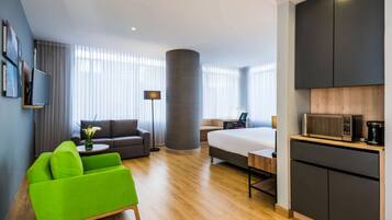 Quarto de hotel moderno com piso de madeira, uma poltrona verde, um sofá, uma cama, uma mesa de escritório, uma TV de tela plana, um abajur e um micro-ondas.