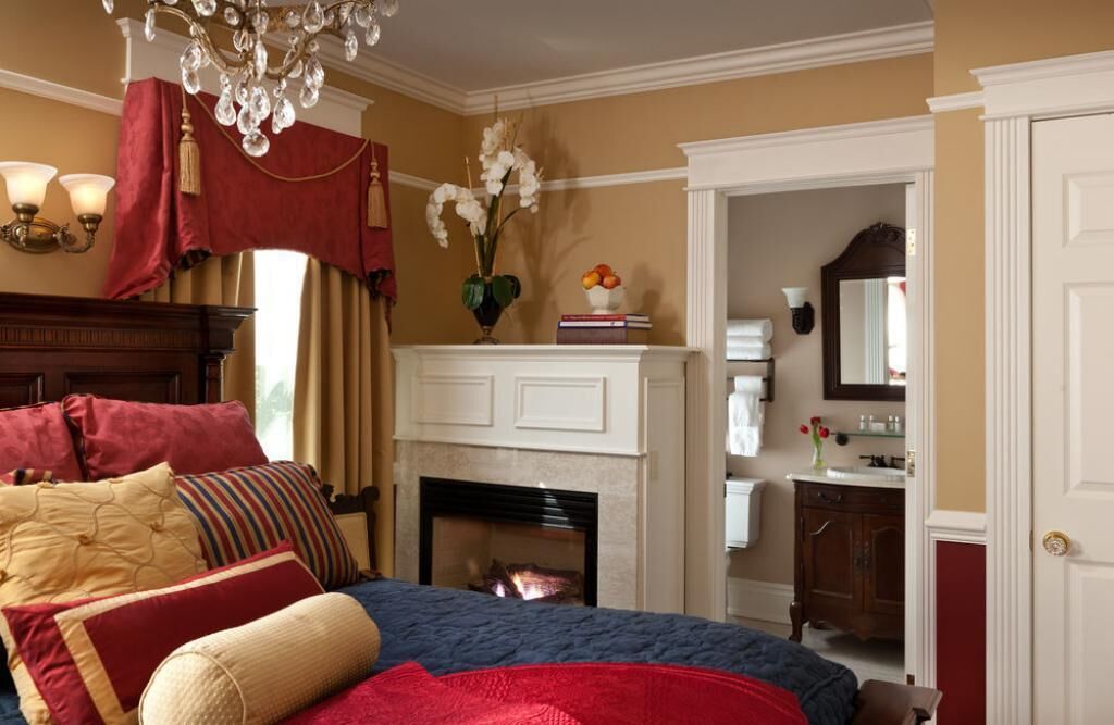 Quarto com lareira, cama com várias almofadas, um candelabro e um espelho no quarto adjacente.