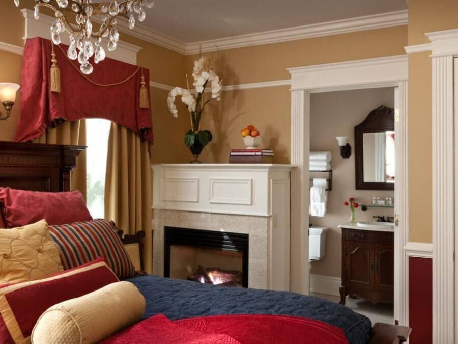 Quarto com lareira, cama com várias almofadas, um candelabro e um espelho no quarto adjacente.