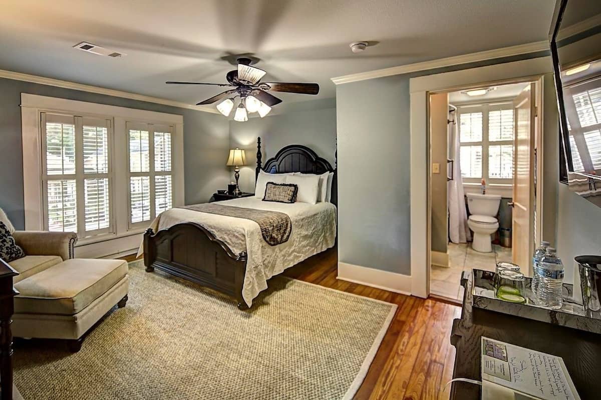 Quarto com cama, escrivaninha, cadeira, ventilador de teto e banheiro visível através de uma porta aberta.