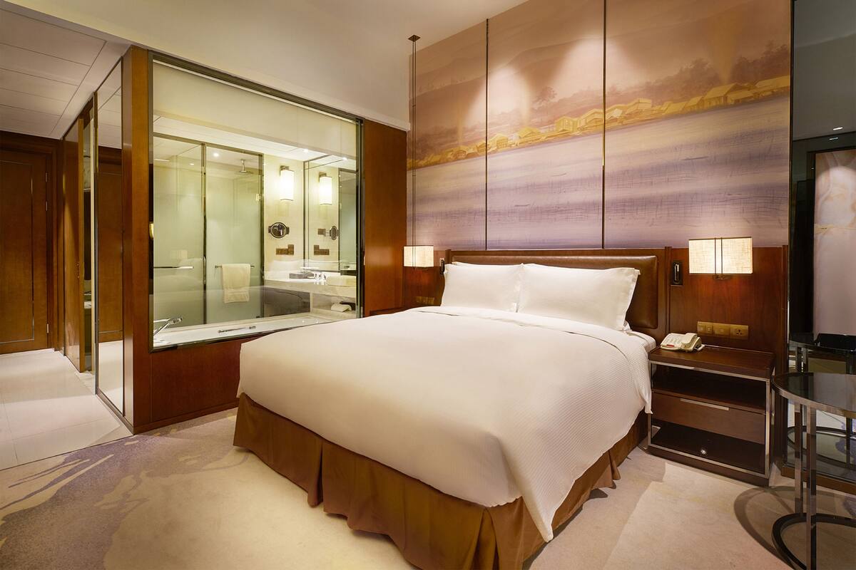 Quarto de hotel com uma cama grande, chuveiro com divisória de vidro, toucador com espelho e televisão fixada na parede.