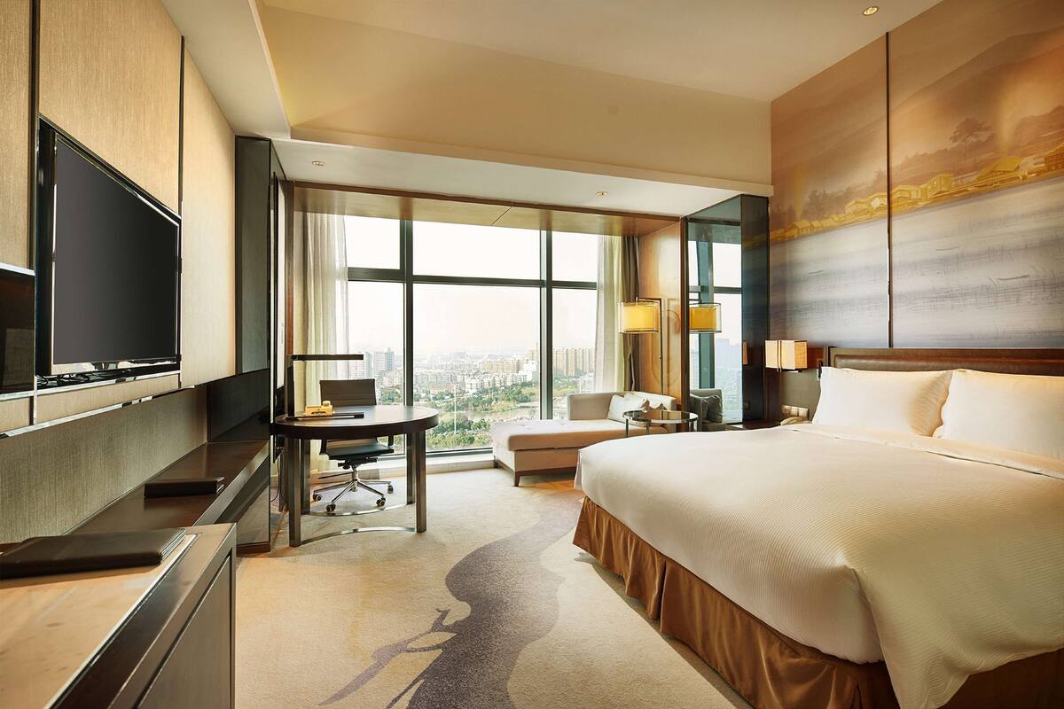 Quarto de hotel com uma cama grande, uma escrivaninha com computador, televisão e vista da cidade através de amplas janelas.