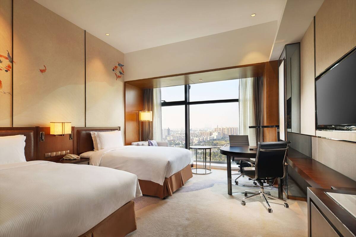 Quarto de hotel com duas camas, uma escrivaninha, uma cadeira, uma televisão e uma janela grande com vista para a cidade.