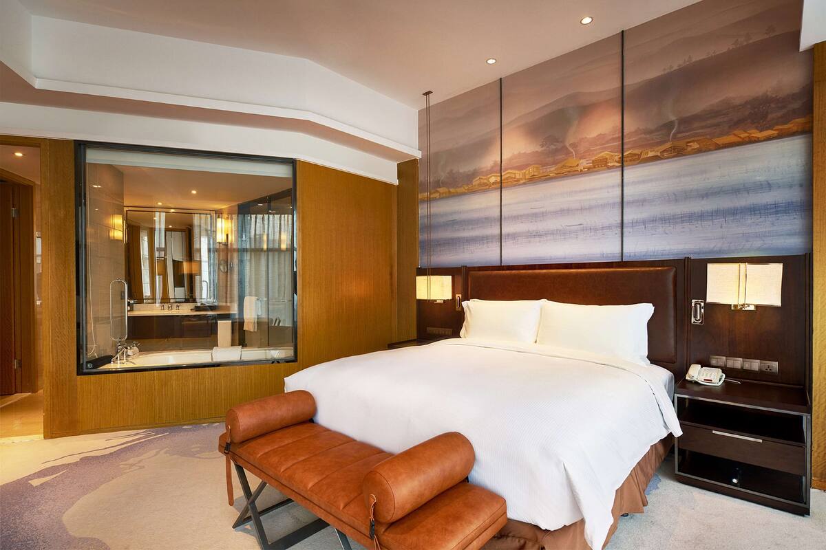 Quarto de hotel moderno com uma cama grande, um banco, casa de banho com paredes de vidro e um quadro grande na parede.