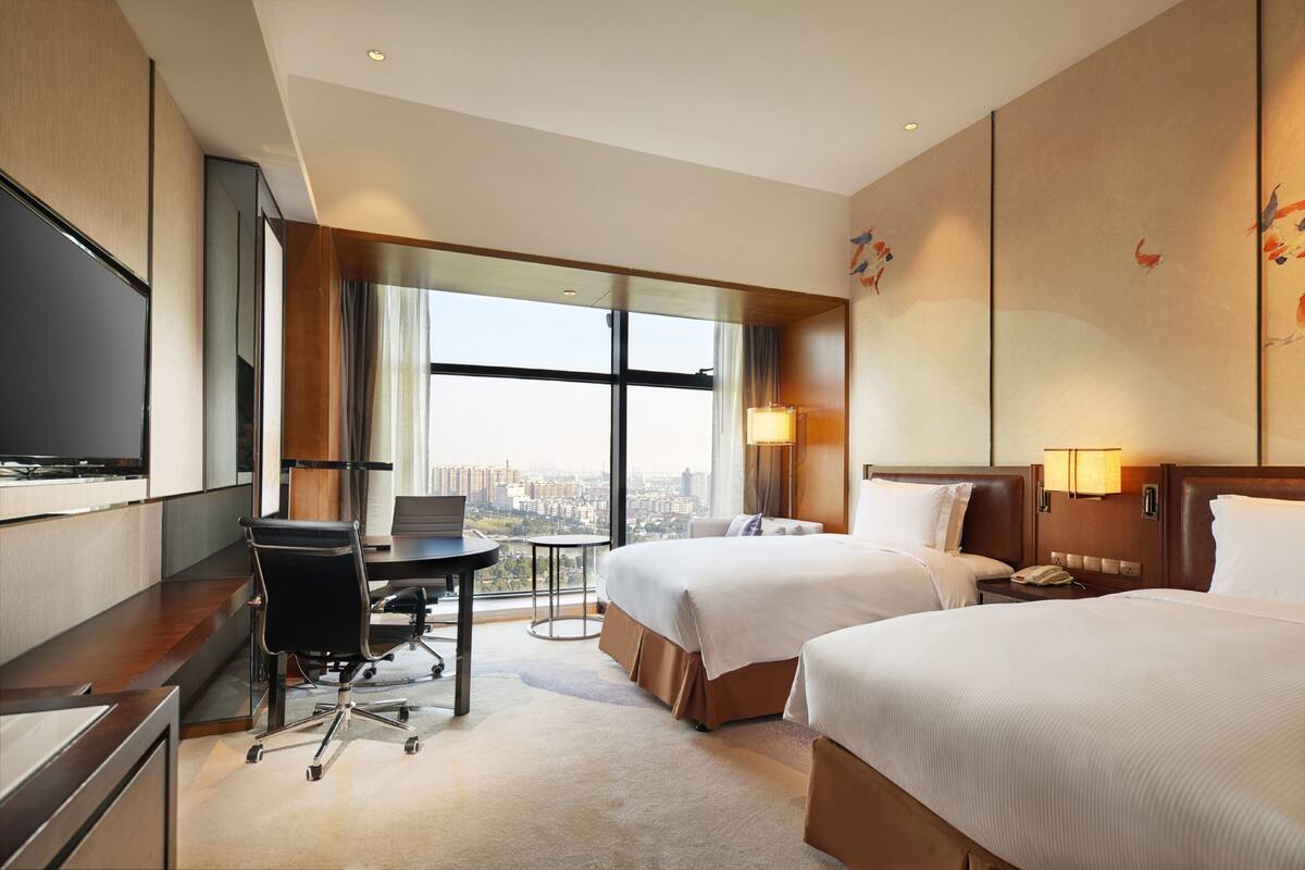 Quarto de hotel com duas camas, uma escrivaninha com cadeira, televisão e vista para a cidade.