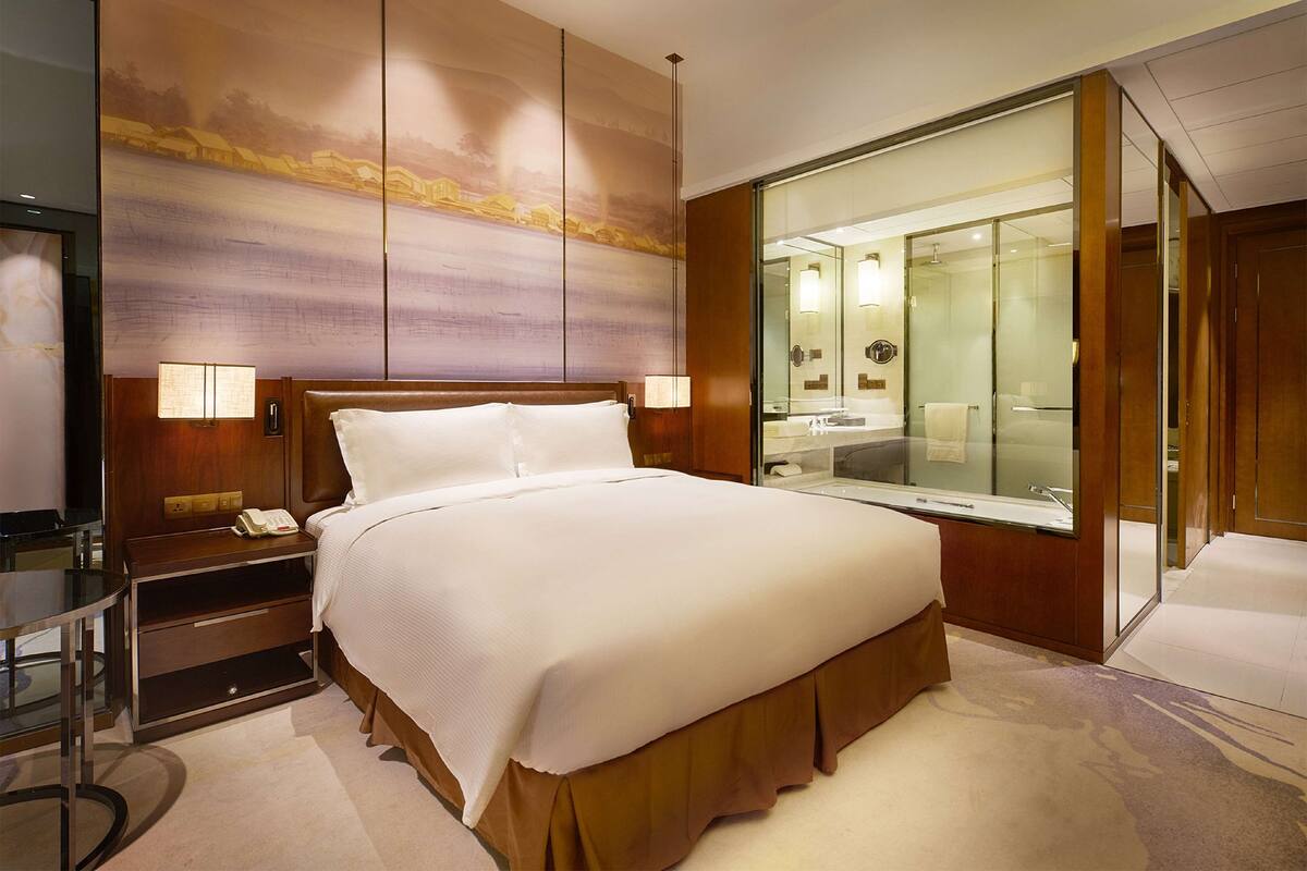 Quarto de hotel com uma cama grande, mesa de cabeceira com abajur, casa de banho com divisórias de vidro e vista para a cidade.