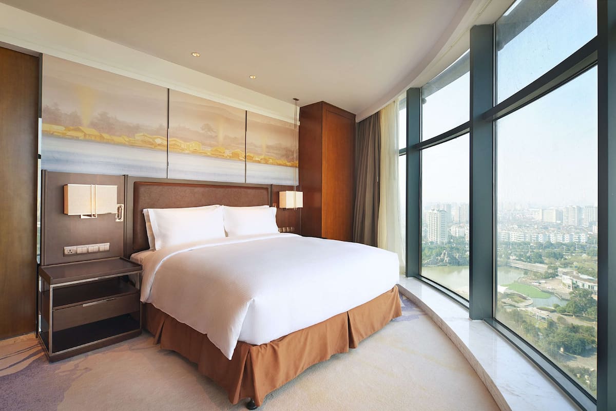 Quarto de hotel moderno com uma cama grande, mesa de cabeceira, luminária na parede e uma janela ampla com vista para a cidade.