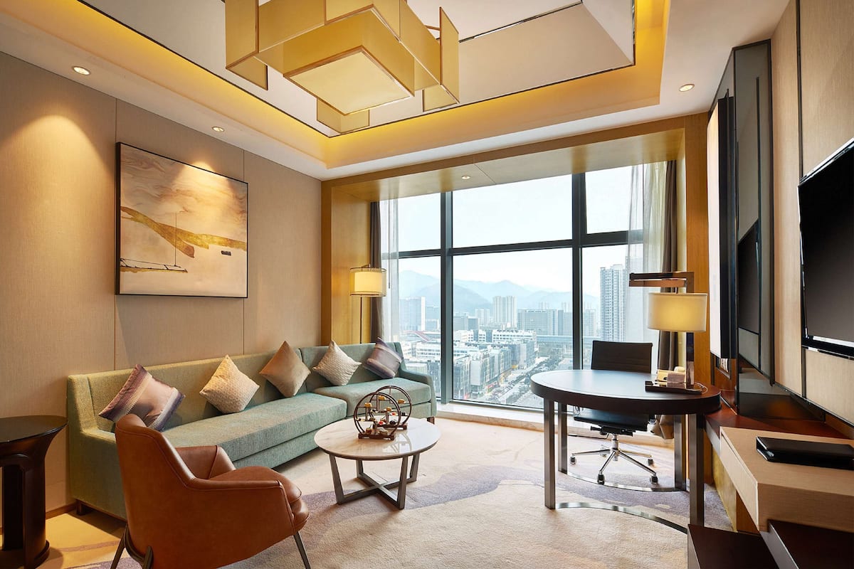 Quarto de hotel moderno com uma janela grande que oferece vista para a cidade, um sofá, uma mesa de centro e uma escrivaninha com cadeira.