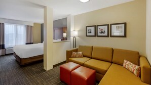 Quarto de hotel com cama, sofá, um banquinho vermelho, luminária e quadros na parede.
