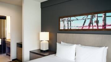 Quarto de hotel com cama, mesa de cabeceira com abajur, quadro na parede e porta à esquerda.