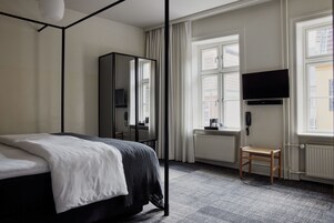 Quarto de hotel moderno com uma cama grande, uma cadeira de madeira pequena, uma televisão fixada na parede e uma janela com cortinas.