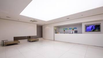 Uma área de recepção moderna, com televisão de tela plana, balcão de atendimento e espaço de espera com bancos.