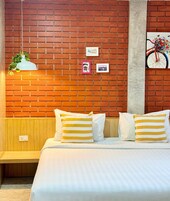 Quarto de hotel com parede de tijolos, cama com almofadas listradas em branco e amarelo, um pequeno banco e luminária fixada na parede.