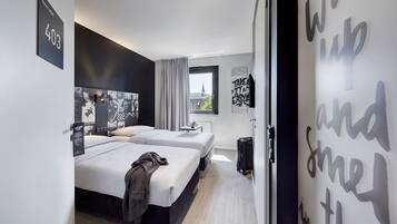 Quarto de hotel moderno com duas camas, janela com vista e um cartaz motivacional na parede.