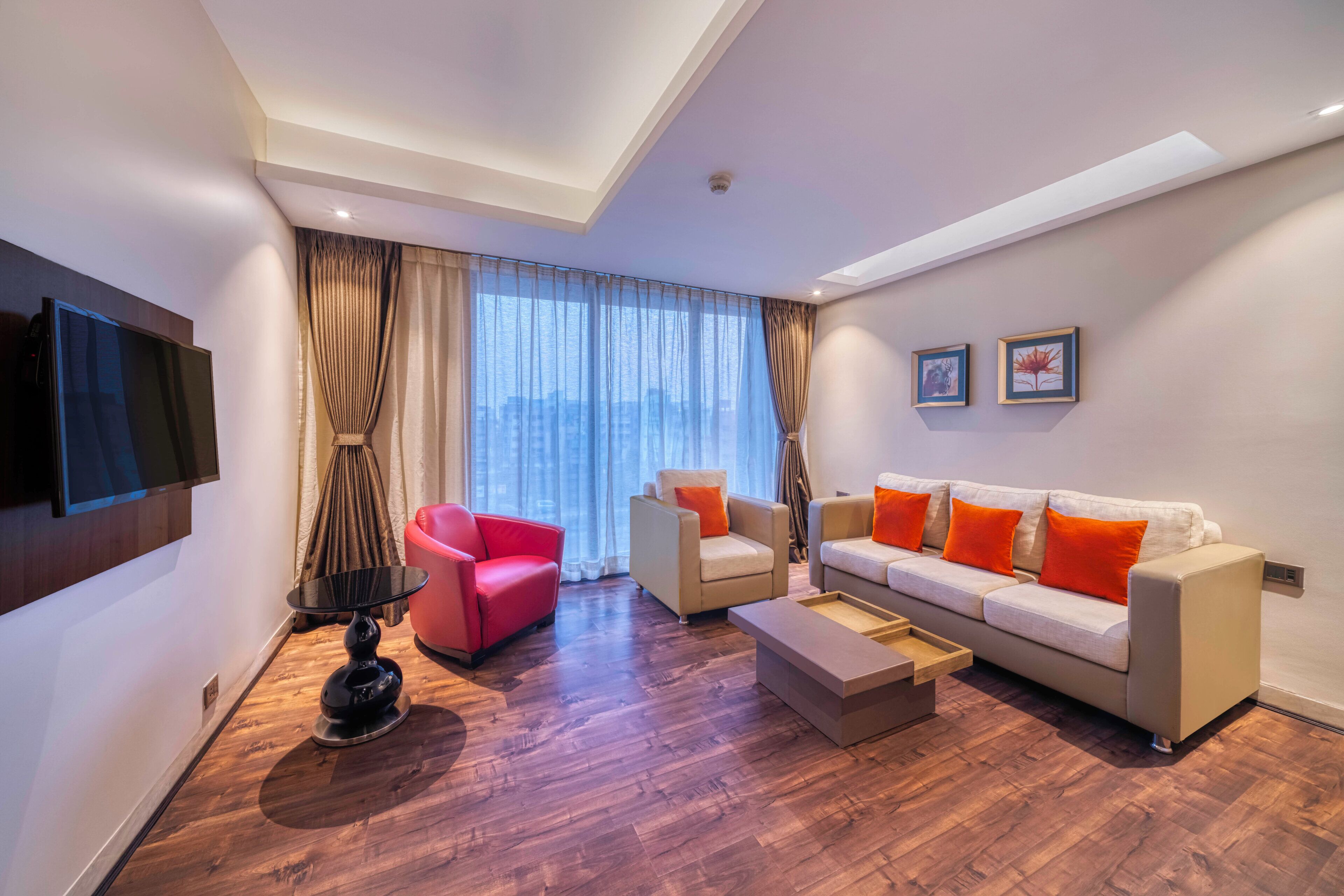 Uma sala de estar moderna com televisão de tela plana, uma poltrona vermelha, um sofá bege com almofadas laranja e uma mesa de centro de madeira.