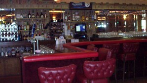 Uma área de bar com banquetas de couro vermelho, um balcão com garrafas e uma tela de televisão.