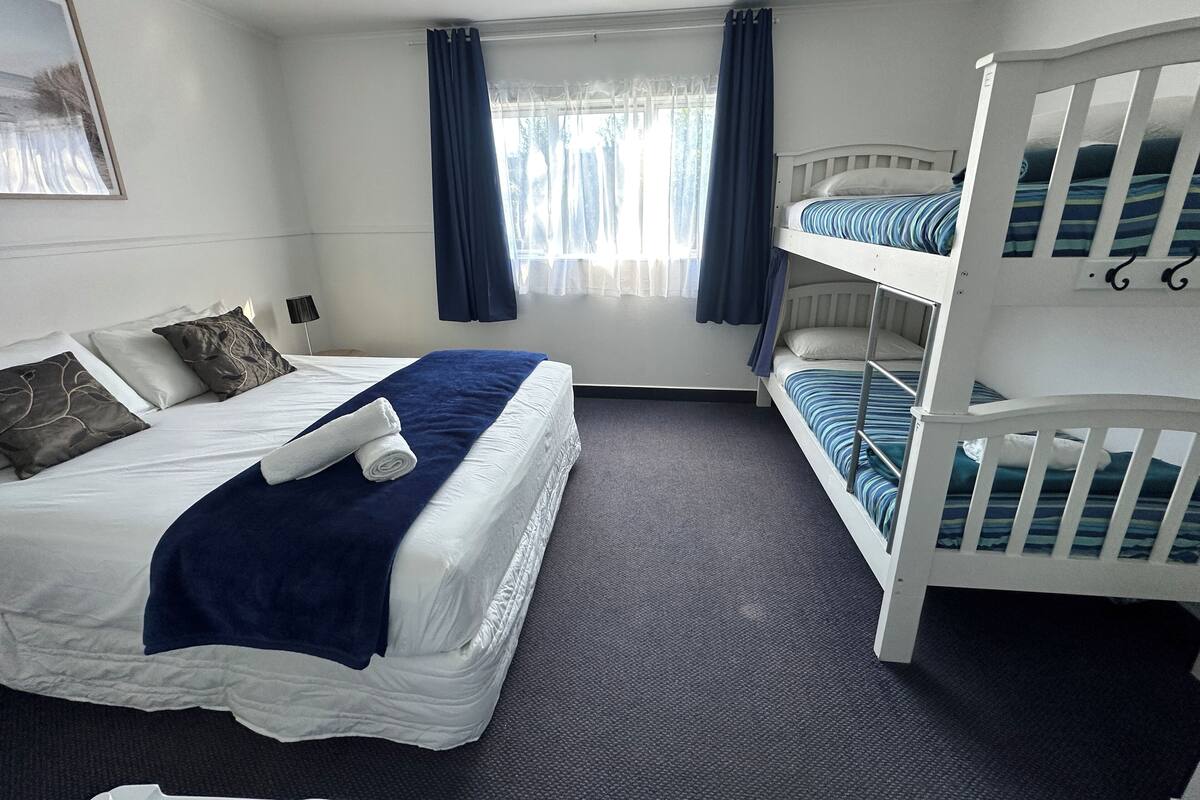 Quarto de hotel com uma cama de solteiro, uma cama beliche, uma janela com cortinas e um quadro na parede.