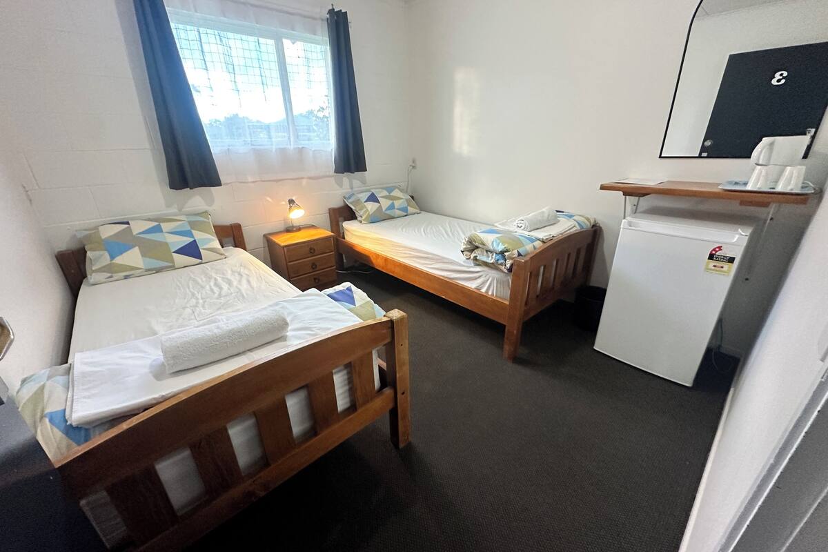 Quarto com duas camas de madeira, uma pequena escrivaninha, um frigorífico e janela com cortinas.