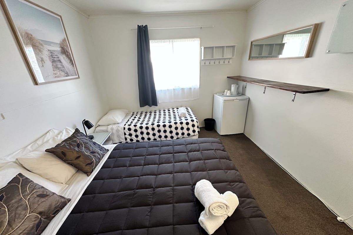Quarto de hotel com uma cama, uma pequena escrivaninha, um espelho, uma prateleira e uma janela com cortinas.