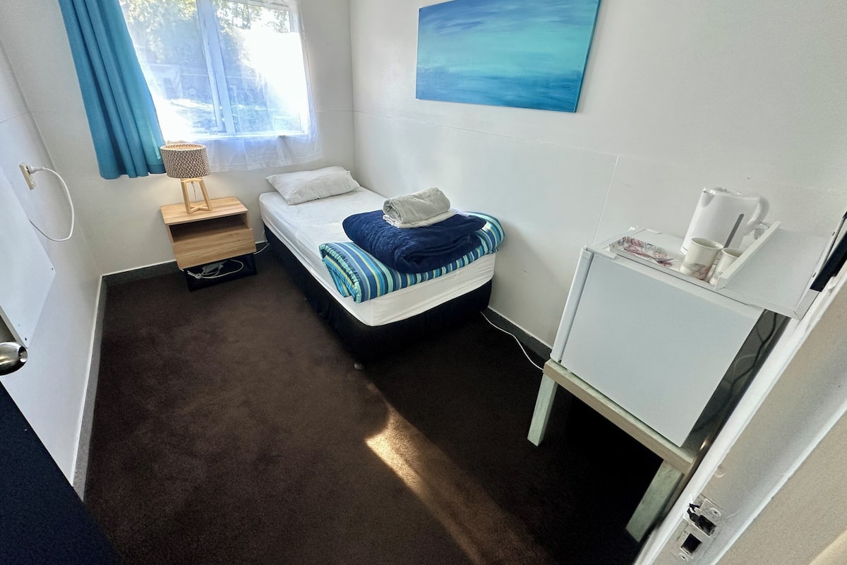 Quarto pequeno e bem iluminado, com uma cama, mesa de cabeceira, abajur e um armário branco com chaleira e xícaras.